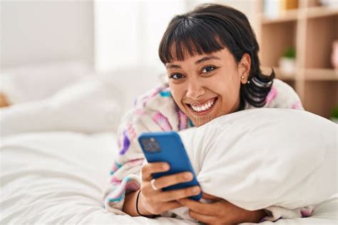 молодая красавица латиноамериканка с смартфоном лежит на кровати в спальне Стоковое Фото