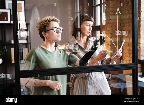 Young Woman With Prosthetic Arm Drawing Graphs On Glass Wall With Her Colleague Examining The