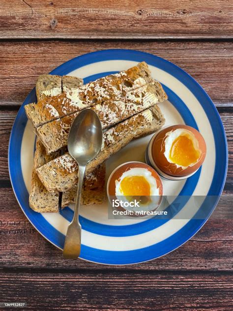 부드러운 삶은 달걀 작은 술 통밀 버터를 구운 빵 조각 콧물 노란색 노른자 나무 곡물 배경 높은 전망을 보여주는 얹은 계란을 얹은 파란색과 흰색 코니쉬 도자기 그릇 차 접시