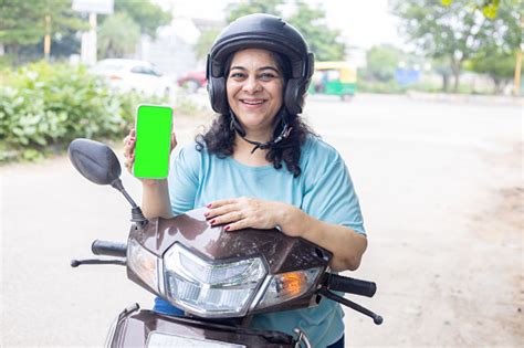 헬멧을 쓴 행복한 노인 인도 여성은 오토바이나 모터 스쿠터에 앉아 광고나 판촉을 하기 위해 빈 디스플레이 화면이 있는 스마트폰을 보여줍니다 오토바이에 대한 스톡 사진 및
