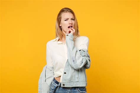 스튜디오에서 밝은 노란색 오렌지 벽 배경에 고립 된 카메라와 갉 아 먹는 손톱을 찾고 데님 캐주얼 옷에 당황 젊은 여자의 초상화 사람들 라이프 스타일 개념입니다 복사 공간을