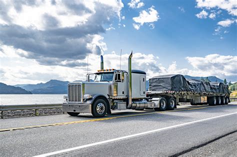 화창한 날에 강을 따라 고속도로에서 운전하는 플랫 베드 세미 트레일러에 덮여있는화물을 운반하는 크롬 부품이있는 클래식 아메리칸 아이돌 빅 리그 화이트 세미 트럭 0명에 대한