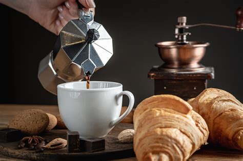 간헐천 커피 메이커의 검은 향기로운 커피를 하얀 컵에 손을 붓고 있습니다 나무 테이블의 머그잔 옆에신선한 크루아상과 커피