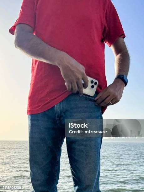 클로즈업 빨간 티셔츠와 데님 청바지의 캐주얼 복장을 입은 알아볼 수 없는 남자의 이미지 스마트폰을 바지 주머니 앞에 놓고 바다 배경 전경에 초점 맞추기 치우기에 대한 스톡
