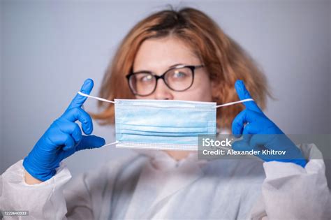 흰색 보호 코트를 입은 젊은 빨간 머리 소녀 의사가 파란색 제리스 장갑을 착용한 그의 얼굴에 의료 용 마스크를 들고 있습니다