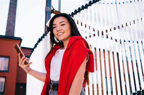 거리 도시성에 서서 카메라를 비웃는 셀룰러 장치를 손에 들고 쾌활한 여성 사용자의 절반 길이 초상화 디지털 스마트 폰을 들고 웃고있는 즐거운 중국 힙스터 소녀 18 19세에