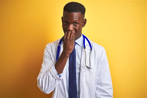 고립 된 노란색 배경 위에 서있는 청진기를 입은 아프리카 계 미국인 의사가 손톱을 물고 손톱을 물고 손으로 스트레스를 받고