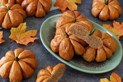 반으로 자른 갓 구운 홈메이드 할로윈 호박 디자인 롤과 타원형 녹색 접시에 녹색 고추 줄기 가짜 가을 잎 녹색 모슬린 회색 배경으로 둘러싸인 살구 잼으로 윤이 나는 빵 빵의