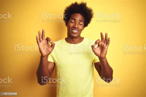 고립 된 노란색 배경 위에 서 있는 티셔츠를 입은 아프리카계 미국인 남성이 손가락으로 명상 제스처를 하고 눈을 감고 미소짓고 있습니다 요가 개념입니다 들이마심에 대한 스톡