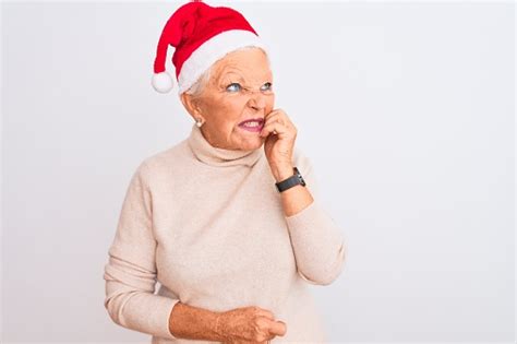 Crhistmas 산타 모자를 쓰고 고립된 흰색 배경 위에 앉아 있는 회색 머리의 수석 여성은 손톱을 물어 뜯는 손톱에 손을 얹고 스트레스를 받고 긴장합니다 불안 문제 12
