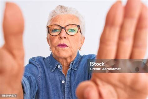 데님 셔츠와 안경을 착용한 회색 머리의 여성은 진지하고 자신감있는 표정 방어 제스처로 정지 표지판을 하는 열린 손으로 고립 된 흰색 배경 위에 셀카를 만듭니다 셀카에 대한