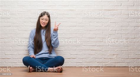 젊은 중국 여자를 표시 하 고 손가락으로 가리키는 벽돌 벽을 통해 바닥에 앉아 자신감과 행복 미소 하는 동안 두 번 가리키기에 대한 스톡 사진 및 기타 이미지 Istock