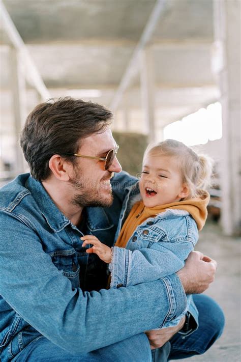 улыбающийся папа обнимает маленькую девочку громко смеясь сидя на корточках возле паддока