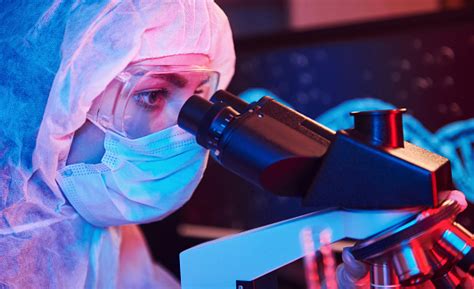 코로나바이러스 백신을 검색하는 컴퓨터 현미경 및 의료 장비가있는 네온 조명 실험실에 앉아 있는 마스크와 흰색 유니폼을 입은