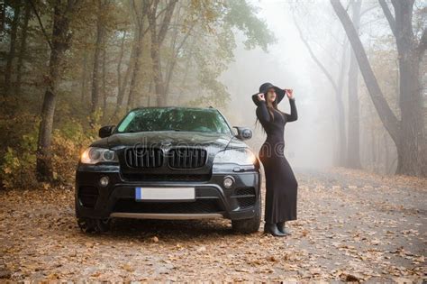 Enigmatic Woman In Black Dress Posing With Suv In A Foggy Forest Stock