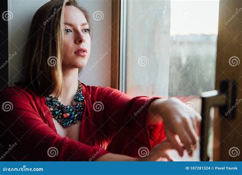 Beautiful Girl In A Red Robe Is Sitting On The Windowsill At Home