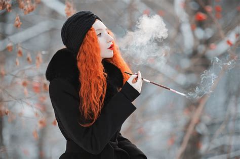 손에서 떠 있는 겨울 숲의 배경에 검은 코트에 붉은 머리를 가진 여자 담배와 그녀의 머리에 터 번으로 밝은 모습으로 빨간 머리