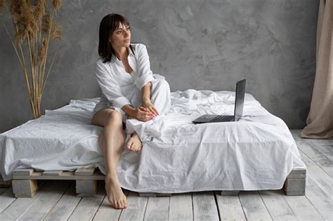 침대에 앉아있는 동안 침실에서 아침에 집에서 노트북으로 일하는 소녀 잠옷에 노트북과 젊은 갈색 머리 온라인과 주말에 원격 작업 사람과 기술 개념 가정의 방에 대한 스톡 사진