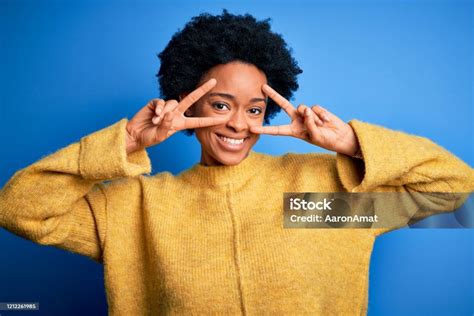 젊은 아름다운 아프리카 계 미국인 아프로 여자 곱슬 머리를 입고 노란 캐주얼 스웨터 얼굴에 손가락으로 평화 기호를하고 승리를