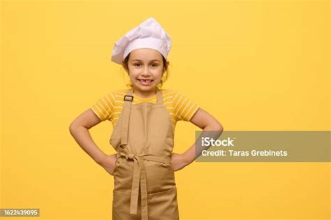 앞치마를 두르고 하얀 요리사 모자를 쓰고 허리에 손을 얹고 카메라를 바라보는 장난꾸러기 소녀 6 7 살에 대한 스톡 사진 및 기타 이미지 Istock
