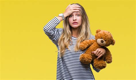 젊은 아름 다운 금발의 여 자가 머리에 손으로 압박 고립 된 배경 위에 테 디 베어 봉 제를 들고 충격 수치심과 깜짝 얼굴로 분노와 좌절 두려워 하 고 실수에 대 한 화가