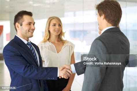 악수하는 사업가들 두 명의 자신감 넘치는 사업가가 배경에 있는 사람들과 함께 사무실에 서서 악수하고 웃고 있다 1개월에 대한 스톡 사진 및 기타 이미지 Istock