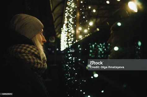 모자를 쓰고 아래 재킷과 볼륨감 있는 스카프를 입은 젊은 금발 여성이 장식된 상점 창문의 화환 카페 클로즈업 창에서 어둠 속에서 보입니다 12월에 대한 스톡 사진 및 기타