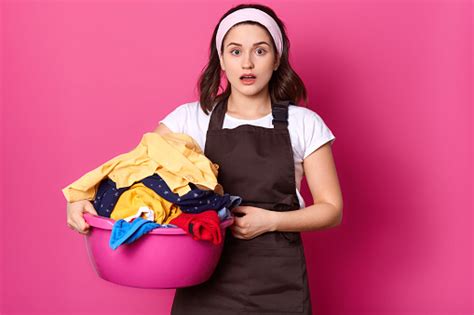 유럽의 주부는 더러운 옷 열린 입으로 스탠드의 전체 분 지를 보유 하 고 밝은 분홍색 배경에 고립 된 포즈 얼굴 표정 충격을 하고있다 가정용 및 국내 집안일 개념입니다