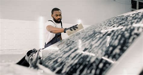 젊은 아프리카 남자 세차 노동자 티셔츠와 작업복을 입고 특수 거품과 스폰지의 도움으로 자동차 앞 유리를 청소합니다 손