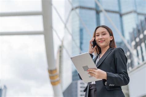 매력적인 젊은 아시아 전문 사업가 숙녀 그녀의 얼굴에 좋은 미소를 지으며 전화로 이야기하면서 방콕시의 중심 비즈니스 지구에있는 디지털 태블릿에 대한 마케팅 및 판매 재고