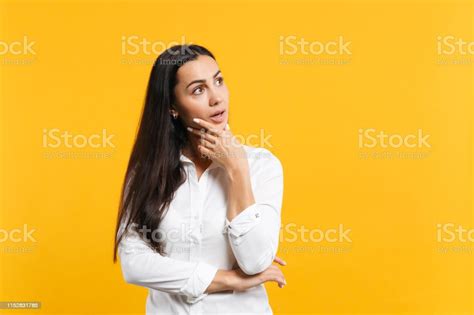 흰색 셔츠에 잠겨있는 매력적인 젊은 여자의 초상화 턱에 손을 넣어 스튜디오에서 노란 오렌지 벽 배경에 고립 된 옆으로 찾고 사람들 라이프 스타일 개념입니다 복사 공간을 모의