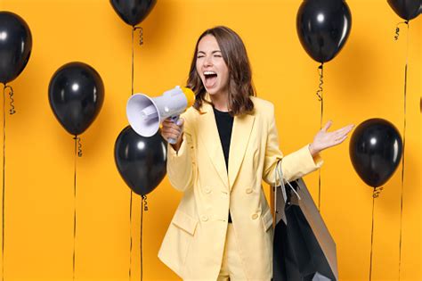 수트 재킷을 입은 미친 젊은 여성은 메가폰에서 비명을 지른 후 공기 풍선 스튜디오 초상화로 노란색 배경에 눈을 감고 쇼핑 후 구매와 패키지 가방을 개최합니다 블랙 프라이데이
