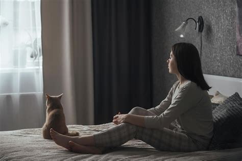 Грустная девушка в пижаме с сезонным аффективным расстройством сидит на кровати и смотрит в окно