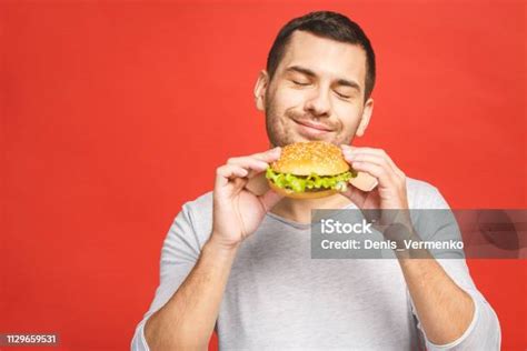 햄버거의 조각을 들고 젊은 남자 학생은 패스트 푸드를 먹는다 햄버거는 도움이 되지 않습니다 매우 배고픈 사람 다이어트 개념입니다 빨간색 배경 위에 고립 컷아웃에 대한 스톡