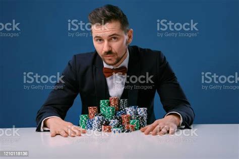 검은 양복을 입은 남자가 파란색 스튜디오 배경에서 포즈를 취하고 있는 컬러 칩더미가 있는 흰색 테이블에 앉아 있다 도박 포커 카지노 클로즈업 갈색 머리에 대한 스톡 사진 및