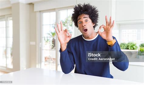 캐주얼 한 스웨터를 입고 집에 앉아있는 젊은 아프리카 계 미국인 남자는 손가락으로 확인 승인 기호를하고 놀랍고 충격을 받았습니다 미친 표현 Omg에 대한 스톡 사진 및 기타