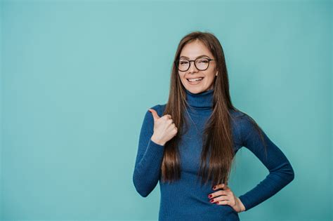 파란색 스웨터와 안경을 쓴 매력적인 이탈리아 젊은 백인 여성이 활짝 웃으며 스튜디오 청록색 배경에 광고를위한 빈 공간이있는 엄지 손가락 제스처를 보여줍니다 모형 쇼핑 판매