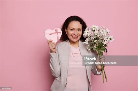 쾌활한 여자는 웃고 통찰력 있는 시선으로 카메라를 바라보며 꽃다발과 하트 모양의 선물 상자를 보여준다 International Womens Day에 대한 스톡 사진 및 기타