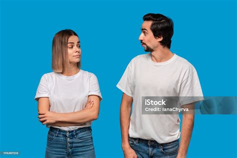 젊은 금발의 여자와 브루넷 수염 남자의 부부는 흰색 티셔츠와 파란색 배경 위에 고립 된 포즈를 서로보고 흰색 티셔츠와 파란색 청바지에 콧수염을 가진 남자 이상적인 부부의