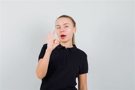 Блондинка показывает знак ок и подмигивает в черной футболке и выглядит оптимистично Бесплатно