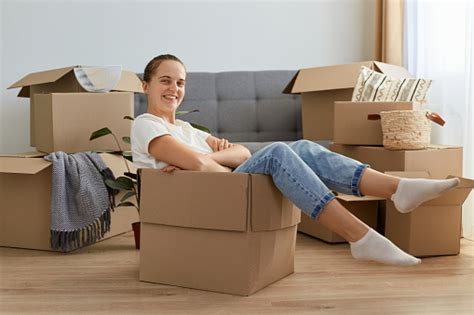 하얀 티셔츠와 청바지를 입고 골판지 상자에 앉아있는 낙관적 인 여성의 실내 샷은 손을 접고 만족스러운 표정으로 카메라를보고 새로운 아파트로 옮겼습니다 30 34세에 대한