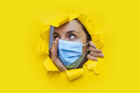 놀란 젊은 여성이 노란 종이에 뚫린 구멍을 들여다보며 의료용 마스크를 고쳐 쓴다 전염병 피로 격리 및 코로나 바이러스 및