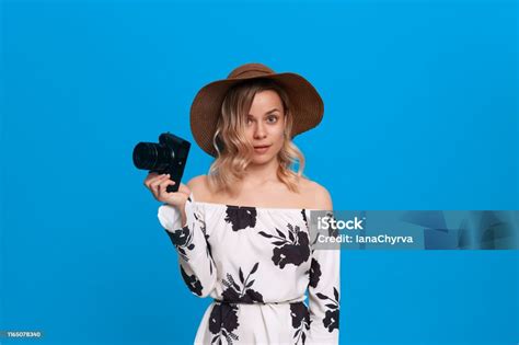 해가 진 모자와 흰색 드레스를 입은 곱슬 금발 머리를 한 모델의 초상화가 파란색 배경에 서있는 복고풍 카메라로 사진을 찍습니다 감정에 대한 스톡 사진 및 기타 이미지