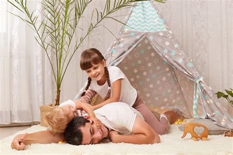 하얀 티셔츠를 입고 아이들과 함께 위그왈 근처 바닥에서 포즈를 취하는 행복한 웃는 남자 아빠와 놀고 웃는 아이들 바닥에 누워 아빠와 즐거운 시간을 보내는 아빠의 실내 촬영