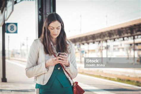 기차역에서 기다리는 동안 스마트 폰을 사용하여 미소 짓는 비즈니스 여성 기차역 플랫폼에 서있는 동안 휴대 전화를 사용하는 매력적인 어린 학생 소녀 Brand Name