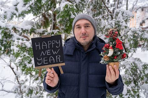 잘 생긴 웃는 남자는 캐주얼 한 옷을 입고 눈 덮인 전나무 근처의 겨울 숲에 서 있습니다 남자는 새해 복 많이 받으세요 비문 소원과 손에 작은 크리스마스 트리 휴일 인사말이