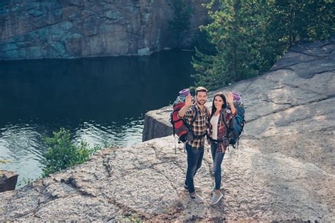 전체 몸 길이 크기 높은 각도 보기 좋은 멋진 배낭 배우자 남편과 귀여운 아내 캐주얼 체크 무늬 셔츠와 청바지를 입고 수염 안녕하세요를 흔들며 물 근처 나무에서 자유 시간을
