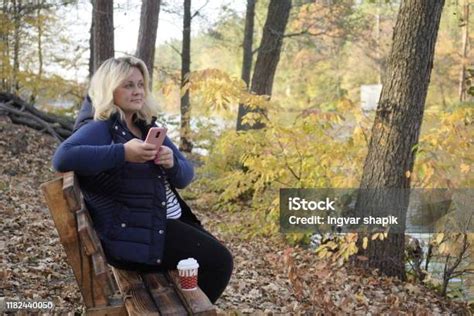 벤치에 앉아 있는 소녀는 호수와 가을 숲을 배경으로 스마트폰을 사용합니다 한 소녀가 호수 앞의 가을 숲의 벤치에 앉아 있다 가을에 대한 스톡 사진 및 기타 이미지 Istock