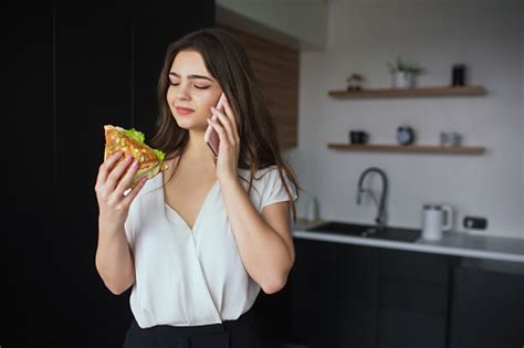 검역 중 부엌에서 젊은 여자 사업가가 전화로 이야기하고 그녀의 손에 맛있는 맛있는 샌드위치를 보세요 원격 작업 중 홈 오피스에서 점심 시간 가구에 대한 스톡 사진 및 기타