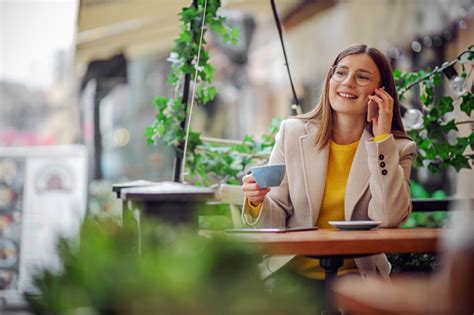 카페 테라스에 앉아 커피 컵을 들고 친구와 전화로 채팅하는 노란색 스웨터에 행복한 미소 갈색 머리 갈색 머리에 대한 스톡 사진